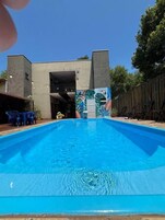 Piscina com superfície de água azul translúcida, cercada por uma vedação de madeira e um edifício com um mural colorido.