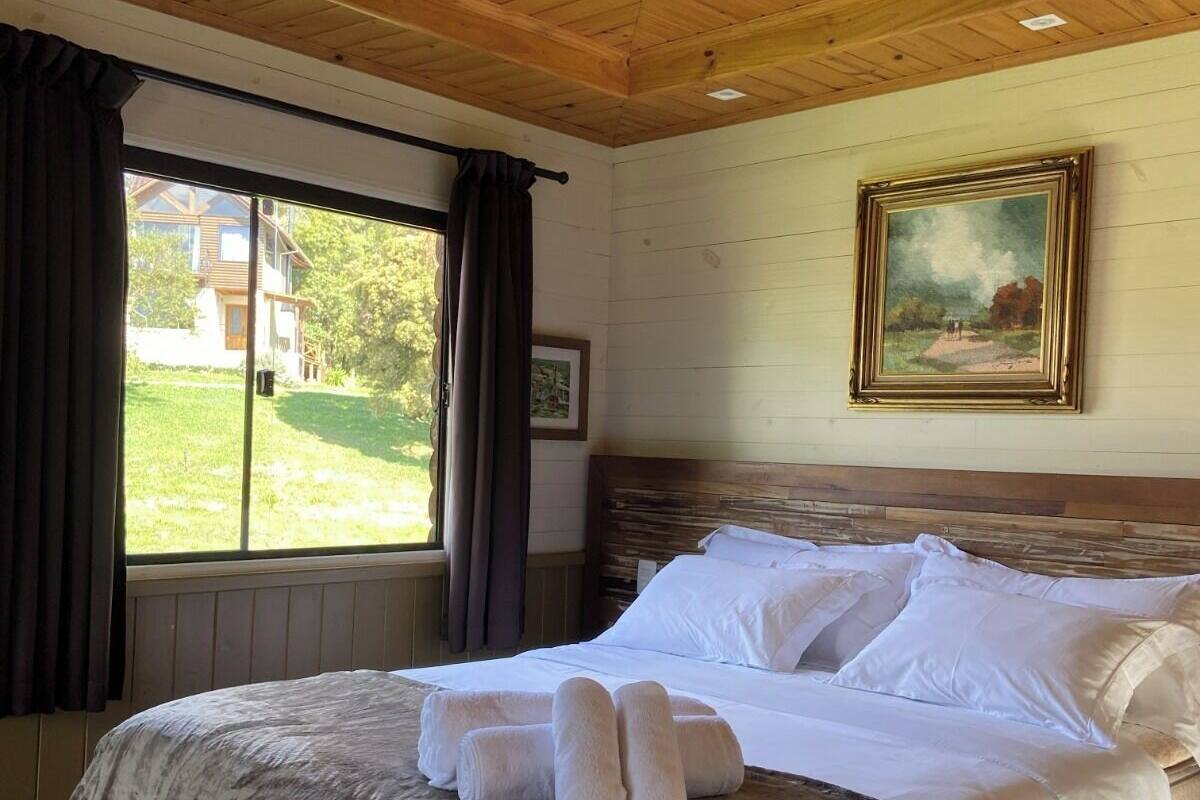 Quarto de hotel com teto de madeira, cama grande com roupa de cama branca, quadro na parede e janela com cortinas.