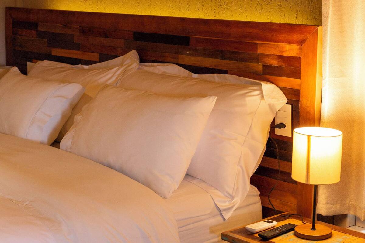Quarto de hotel com cabeceira de madeira, cama com roupa de cama branca, abajur e controle remoto na mesa de cabeceira.