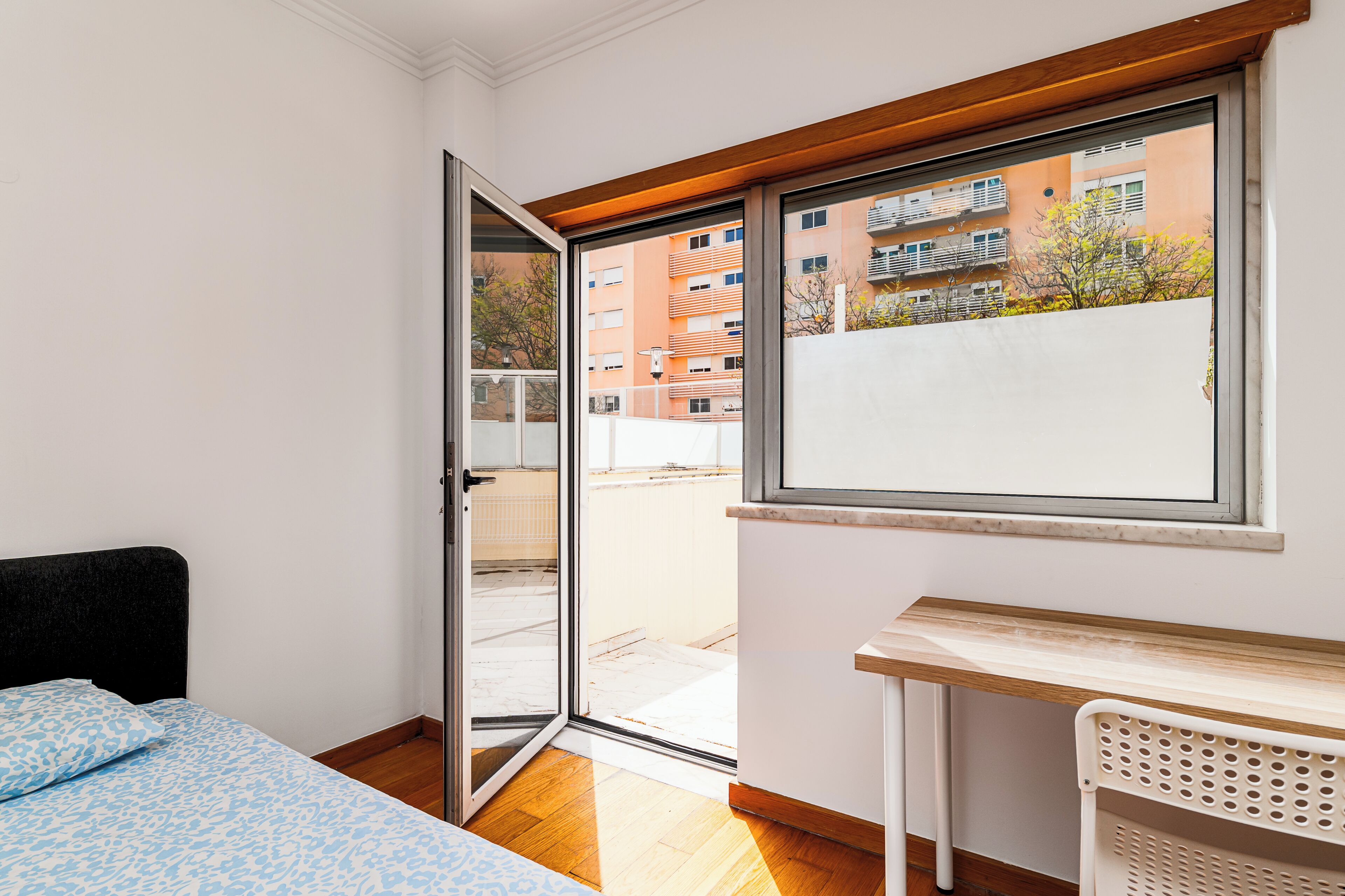 Quarto com cama, escrivaninha, janela com vista para edifícios e porta que dá para o exterior.