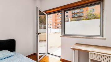 Quarto com cama, escrivaninha, janela com vista para edifícios e porta que dá para o exterior.