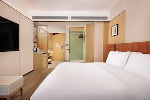 Quarto de hotel com cama, escrivaninha, espelho, televisão, guarda-roupa e chuveiro.