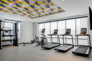 Academia com esteiras, bicicletas ergométricas e teto com padrão geométrico colorido.
