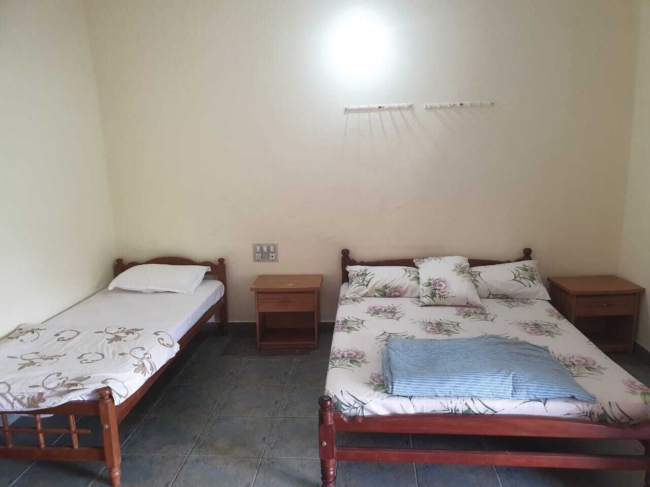 Quarto com duas camas de madeira, cada uma com uma mesa de cabeceira. As camas têm roupa de cama florida, e há um candeeiro na parede.