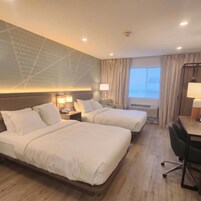 Quarto de hotel com duas camas, uma escrivaninha, uma cadeira, um abajur e uma janela com cortinas.