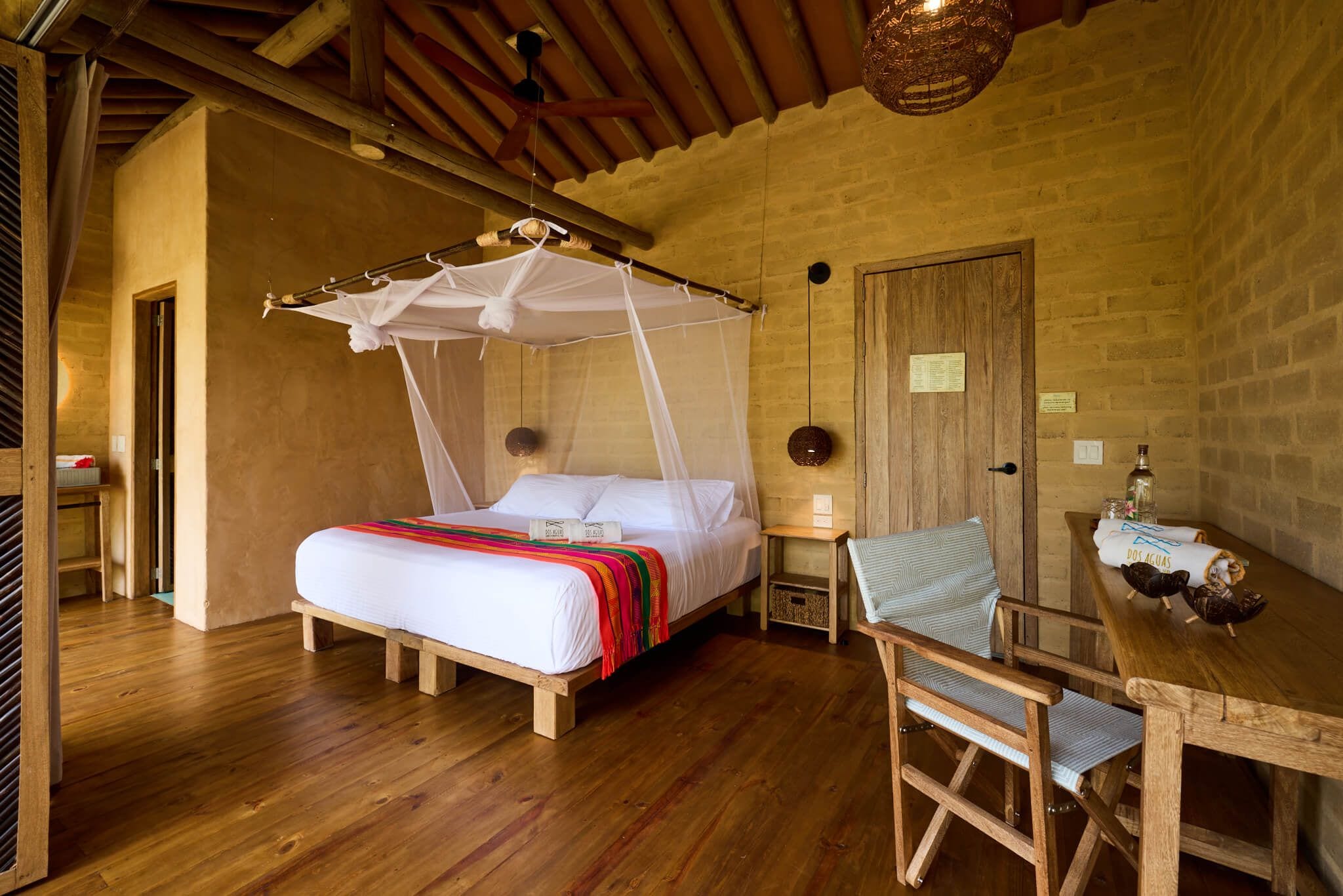 Quarto com estrutura de madeira, cama sob um dossel, mesa de madeira com cadeiras e parede de tijolos.