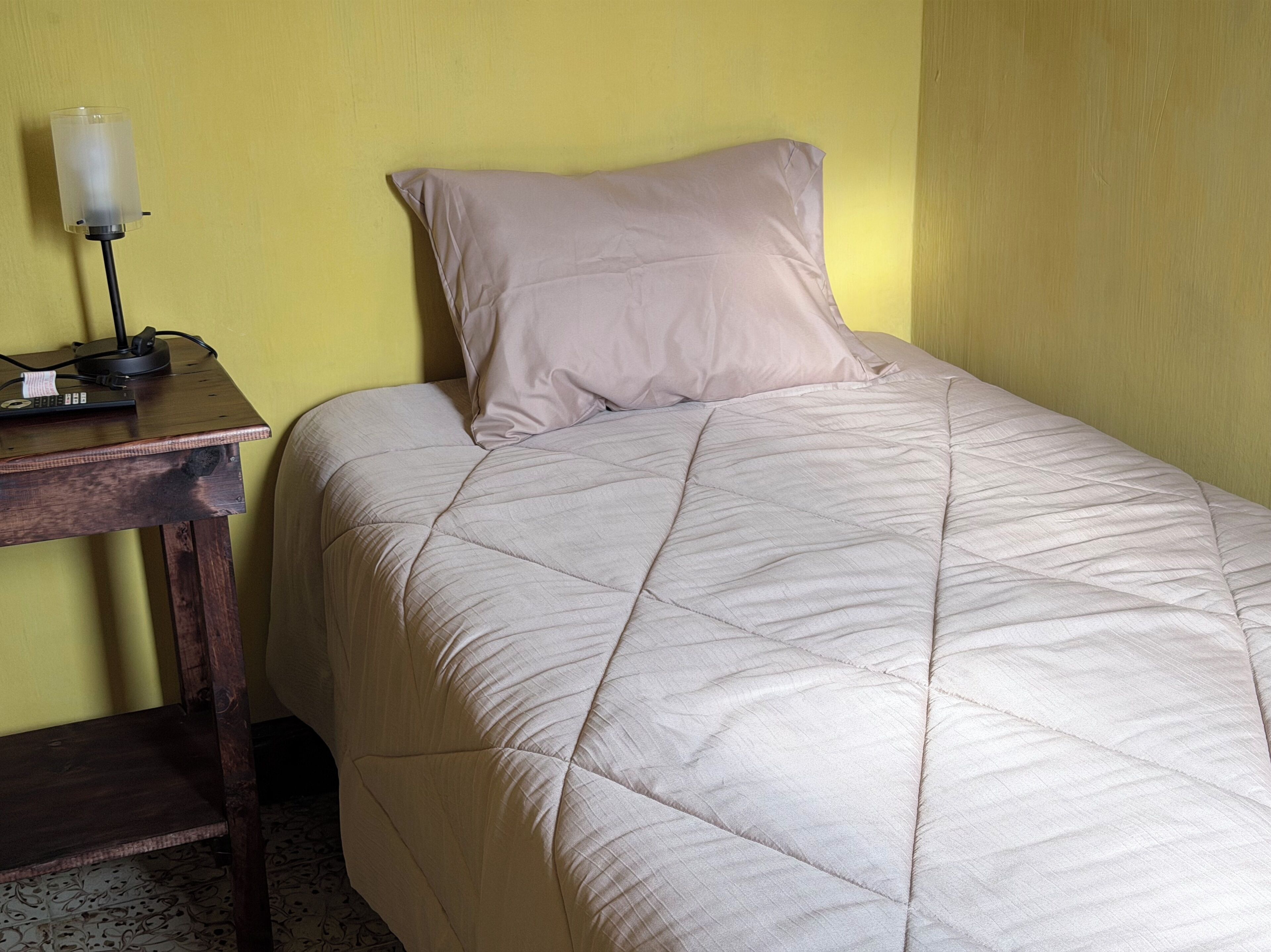 Uma cama de solteiro com cobertura acolchoada, uma mesa de cabeceira com abajur e uma régua de tomadas.
