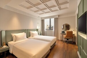 Quarto de hotel com duas camas, uma escrivaninha, uma cadeira, uma janela grande e vista para a cidade.