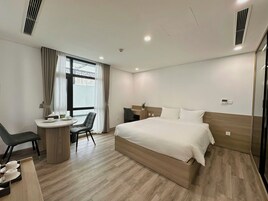 Quarto de hotel com cama, escrivaninha com cadeira, mesa pequena e janela com cortinas.