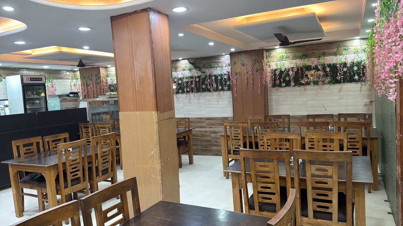 Interior de restaurante com mesas e cadeiras de madeira, um balcão e plantas decorativas nas paredes.