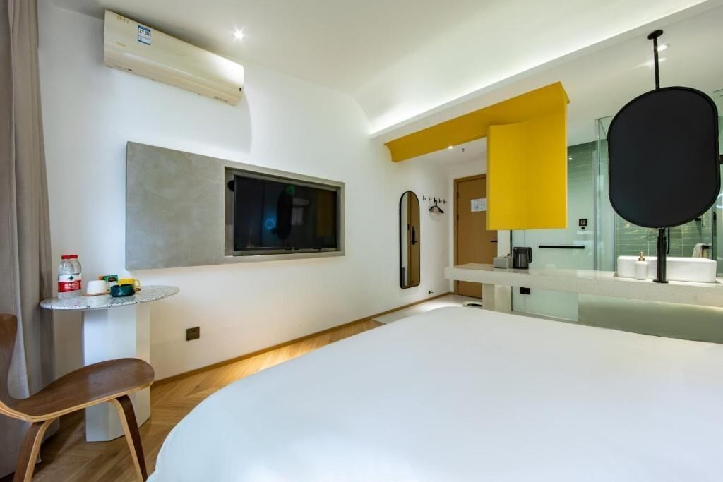 Quarto de hotel moderno com uma cama grande, uma mesinha redonda com uma cadeira, televisão, espelho e banheiro com pia e vaso sanitário.