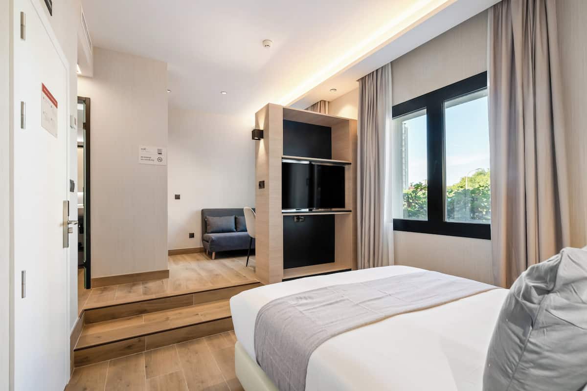 Quarto de hotel moderno com cama, área de estar com poltrona, televisão embutida e janela com cortinas.