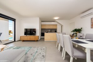 Uma sala de estar moderna com televisão, mesa de jantar e cadeiras, uma cozinha com armários de madeira e uma porta de vidro deslizante que dá para uma área externa.