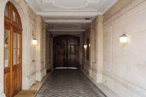Um corredor com uma grande porta de madeira, duas luminárias na parede e chão ladrilhado.