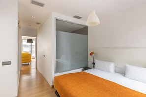 Quarto de hotel moderno com cama grande, divisória de vidro, poltrona amarelo vibrante e vista para a cidade.