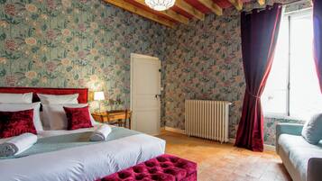 Quarto com cama, um banco estofado vermelho, janela com cortinas, radiador e teto de madeira.