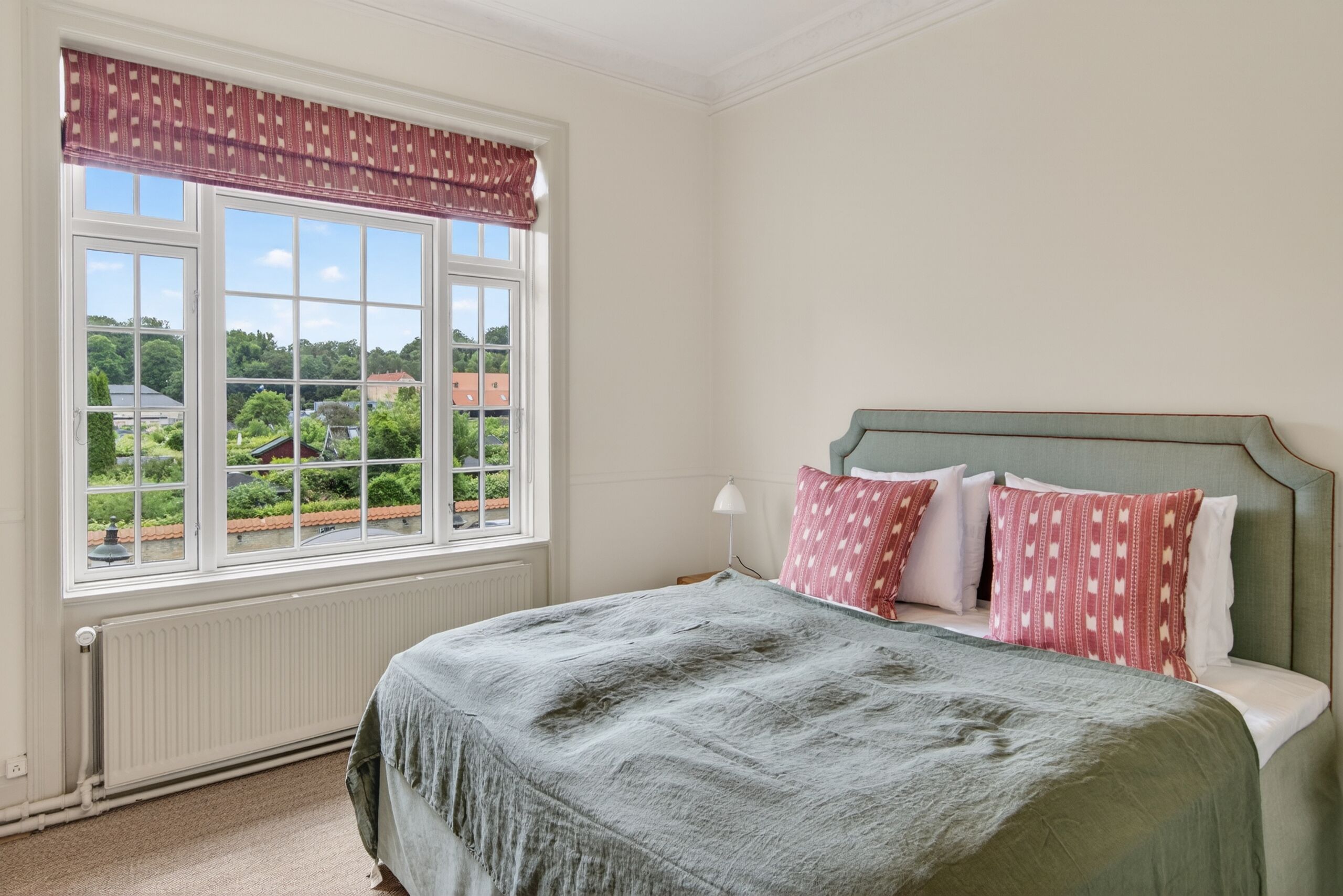 Quarto com cama, janela com vista para área verde e edifícios, e radiador.