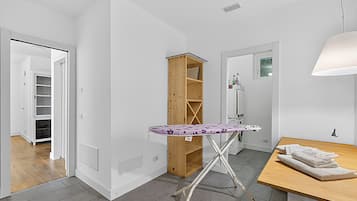 Quarto com tábua de passar roupa de madeira, estante de madeira, mesa com toalha dobrada e porta que dá para outro cômodo.