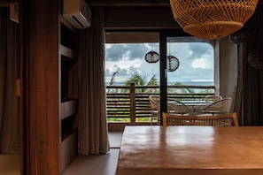 Quarto com mesa de madeira, cadeiras de vime e vista para o oceano através de uma grande janela.