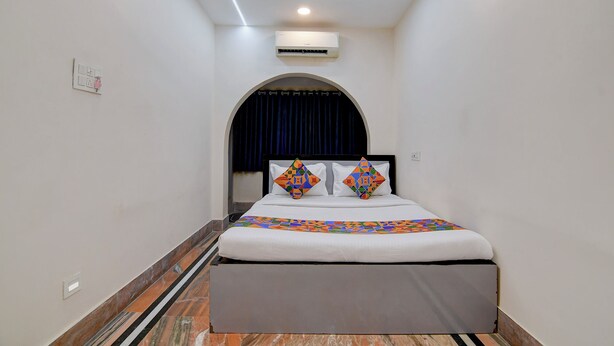 Quarto de hotel estreito com cama, unidade de ar condicionado e pequena janela com cortina.