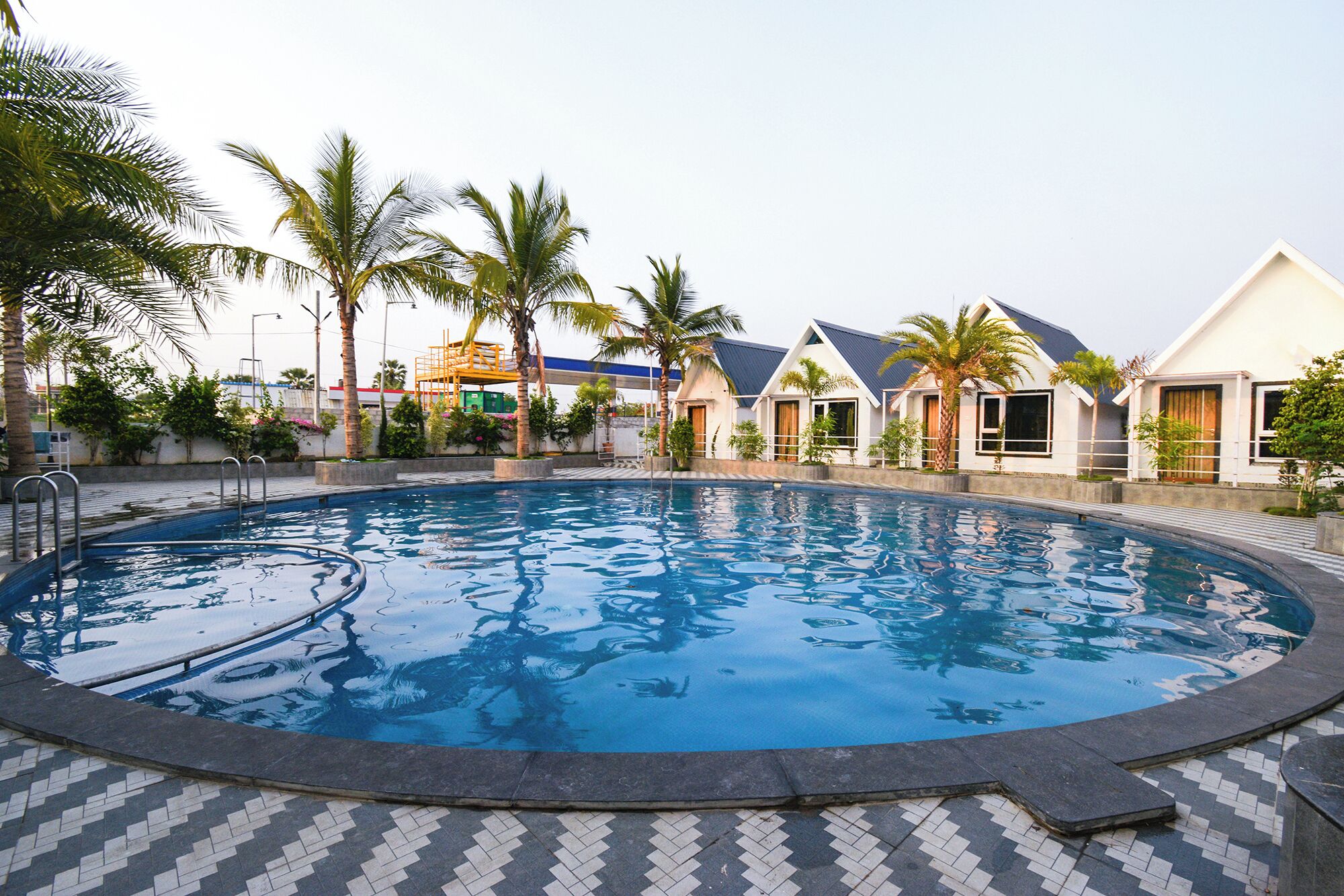 Uma piscina com um deck ladrilhado, rodeada por palmeiras e uma fileira de casas com telhado azul.