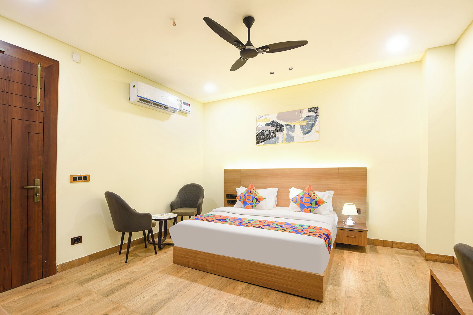 Quarto de hotel com cama, ventoinha no teto, aparelho de ar condicionado, pequena mesa, duas cadeiras e mesa de cabeceira com luminária.