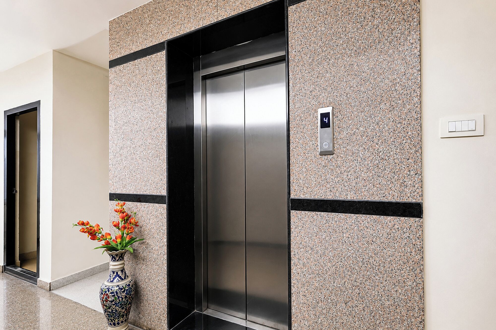 Um elevador com visor digital, parede com padrão salpicado e um vaso com flores.