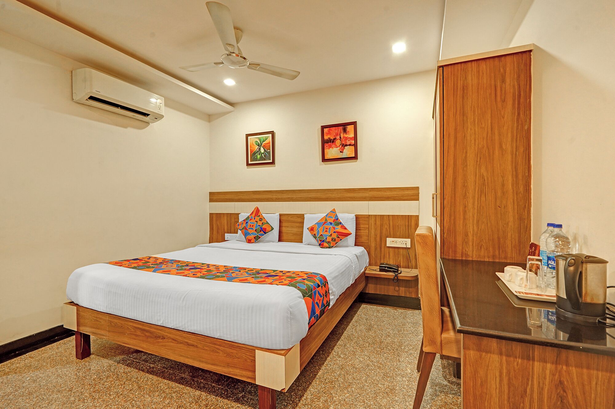 Quarto de hotel com cama, escrivaninha, cadeira, ar condicionado e duas imagens emolduradas na parede.
