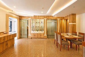 Quarto de hotel moderno com um armário de madeira, porta de vidro, área de jantar com mesa e cadeiras, e um espaço para lavar as mãos.