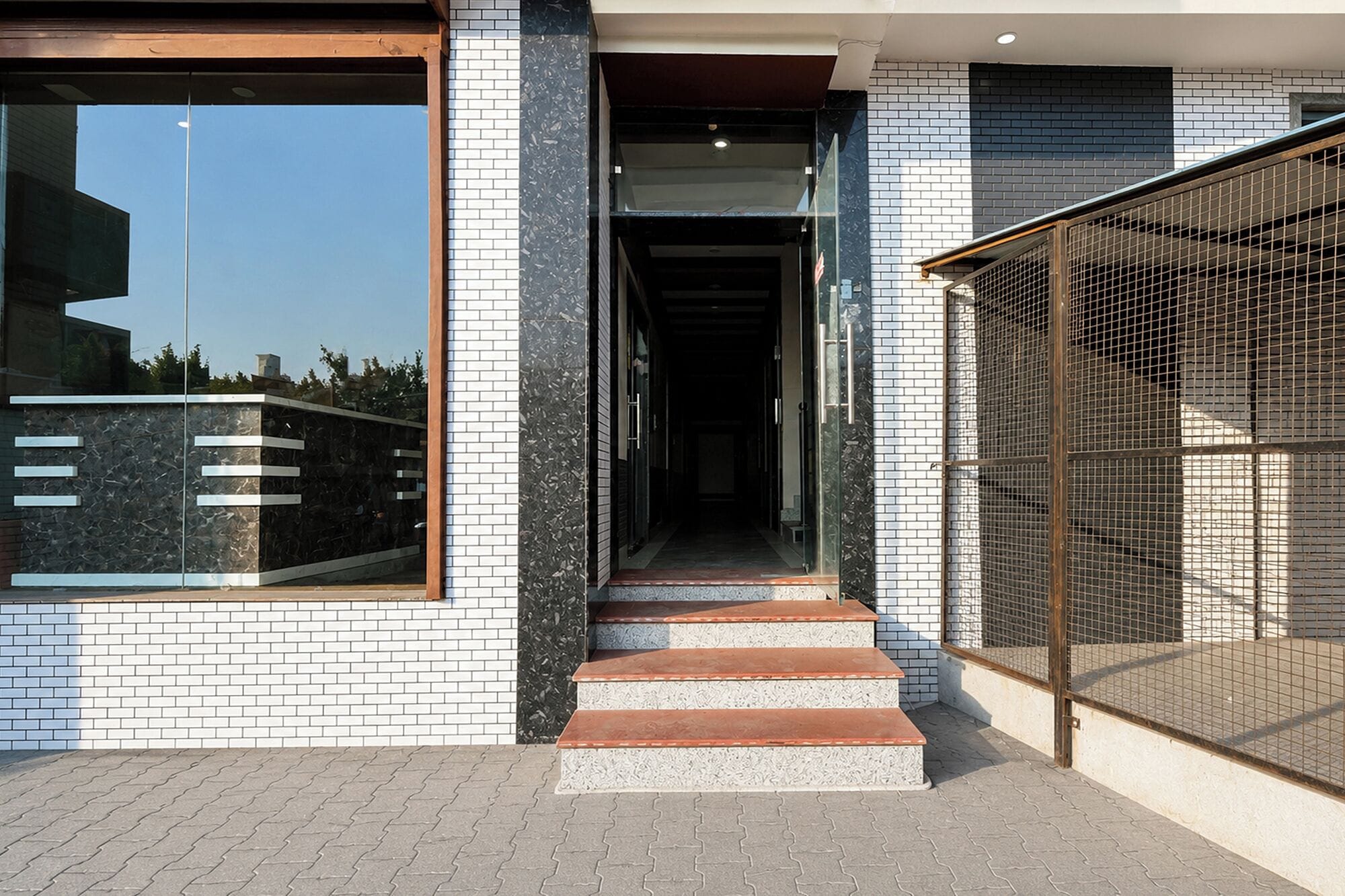 Entrada de um edifício moderno com porta de vidro, portão metálico e escadaria que dá acesso ao interior.