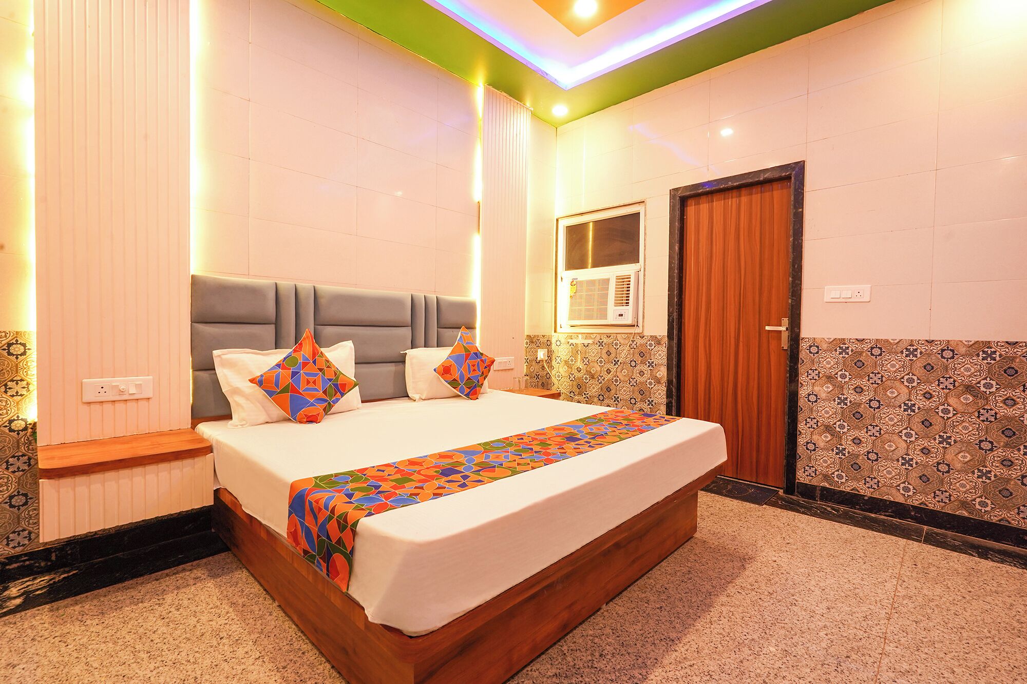 Quarto de hotel com cama, mesa de cabeceira de madeira, janela com ar condicionado, porta e paredes com padrão.