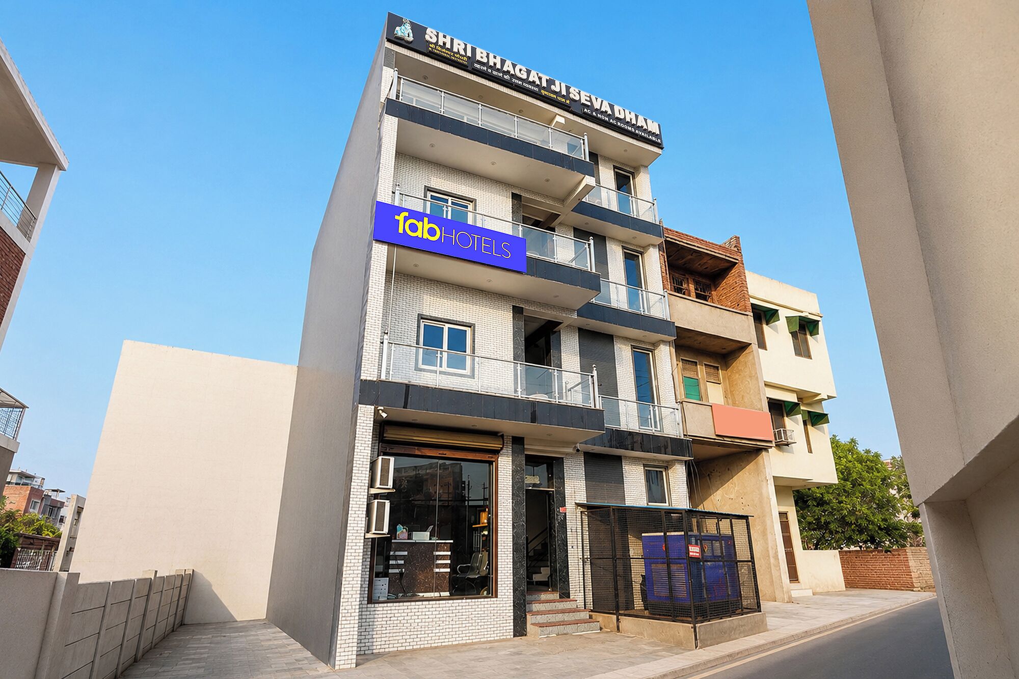 Um edifício de vários andares com uma placa indicando 'SHRI BHAGAT JISEVA DHAM' e uma placa 'fabHOTELS' indicando um hotel.