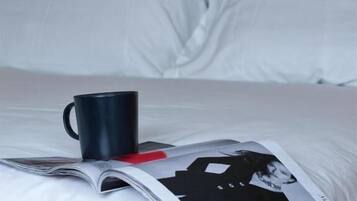 Uma cama com lençóis brancos, uma caneca preta e uma revista com capa em preto e branco.