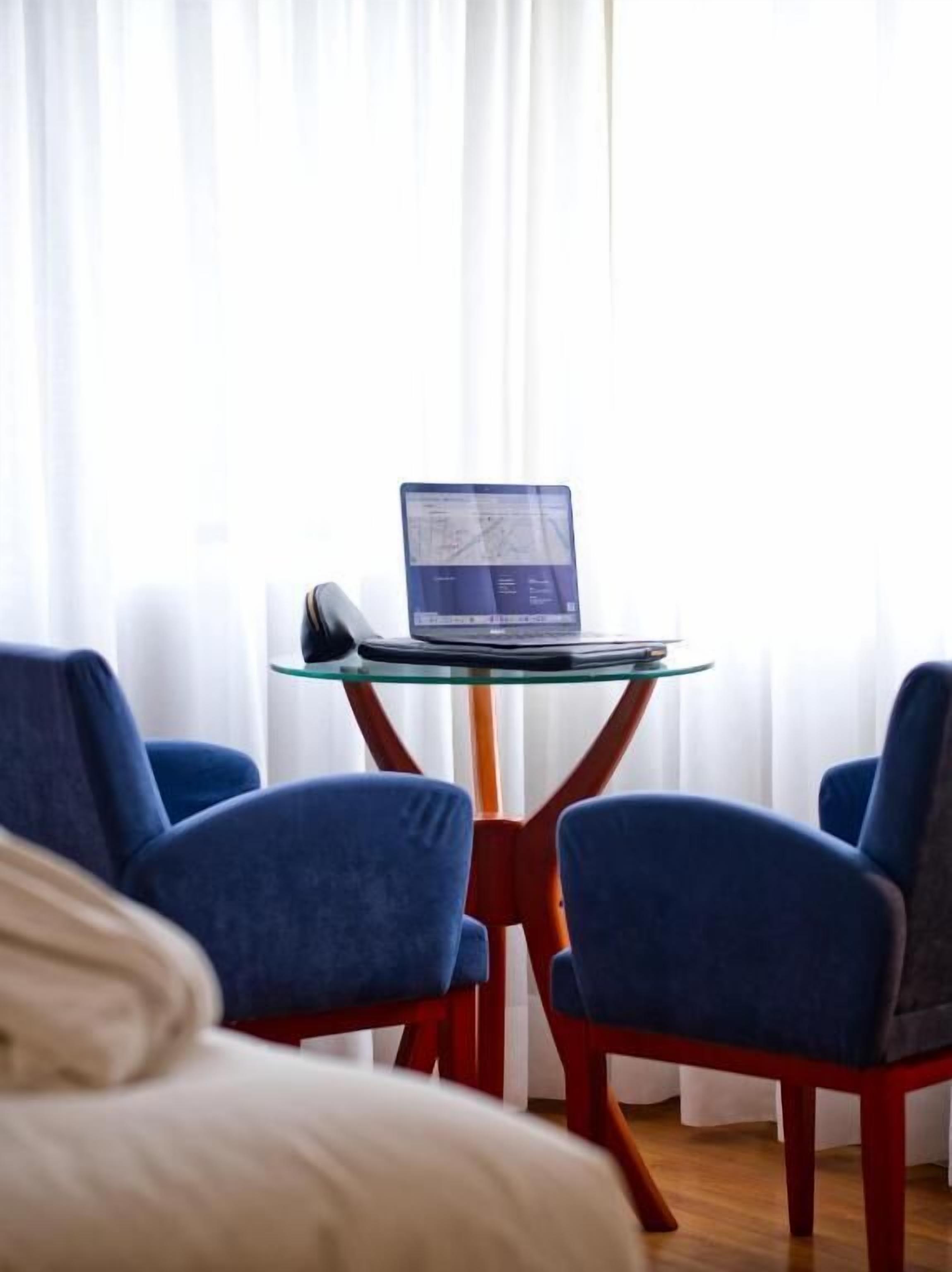 Quarto com mesa de vidro, um laptop e cadeiras azuis.