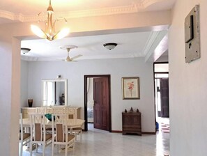 Uma sala de jantar com mesa e cadeiras, um ventilador de teto, um candelabro e uma porta que dá para outro cômodo.