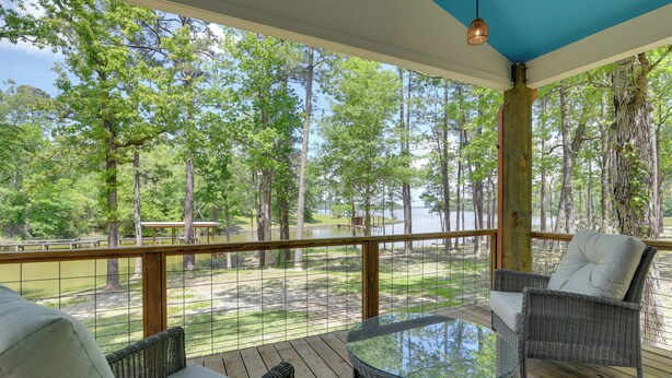 Um deck coberto com móveis de vime, uma mesa de vidro e vista para um lago rodeado de árvores.