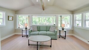 Quarto espaçoso com um sofá verde, duas mesas laterais, uma mesa de centro redonda e grandes janelas com vista para árvores.