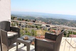 Varanda com mobiliário de vime, uma mesa pequena e vista para uma cidade e colinas.