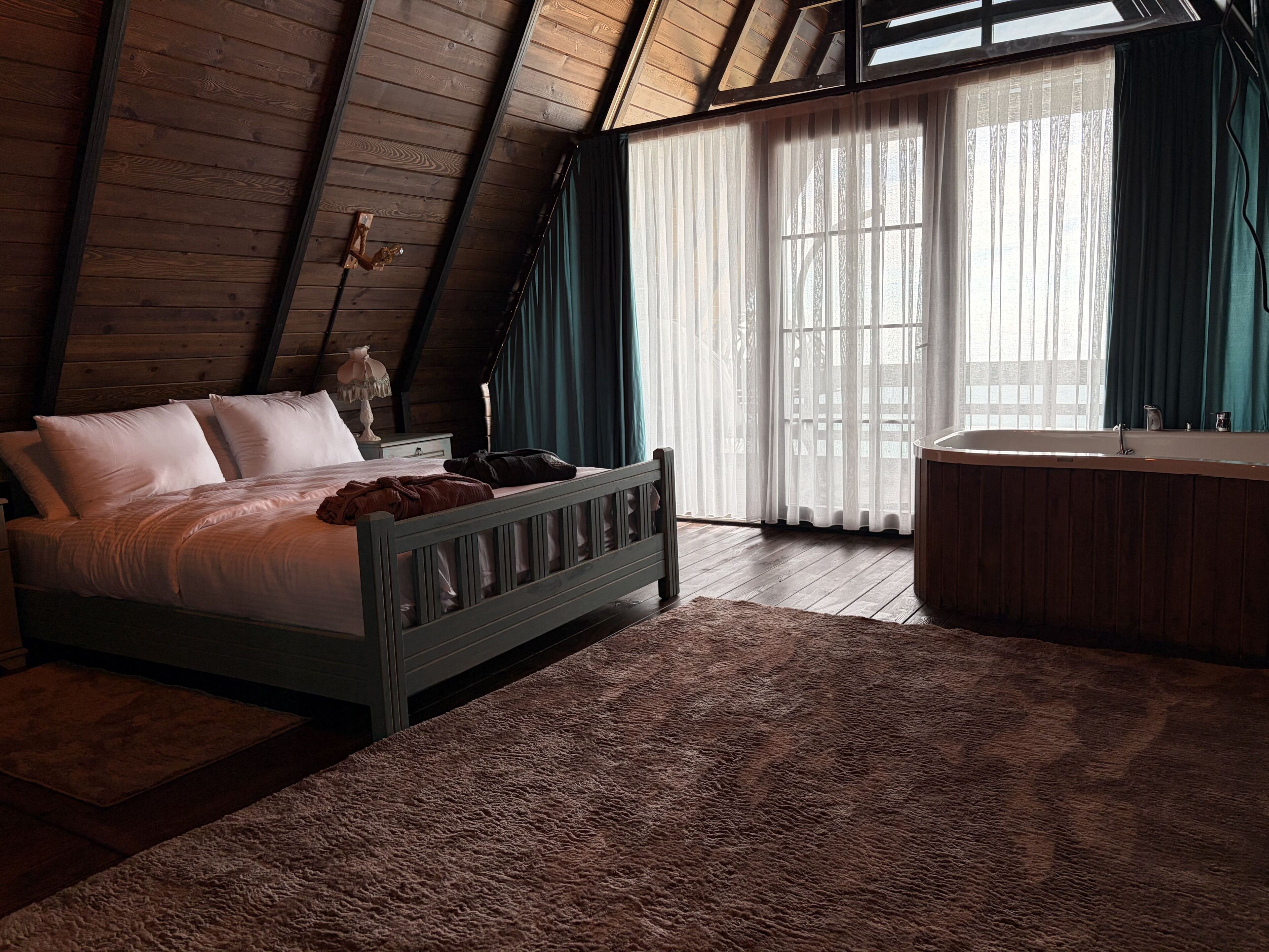 Quarto com cama de madeira, uma janela grande com cortinas, piso de madeira e uma banheira.