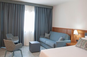 Quarto de hotel com cama, sofá, cadeira, um pequeno banquinho, mesa de cabeceira com abajur e janela com cortinas.