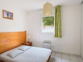 Quarto com cabeceira de madeira, cama com roupa de cama branca, mesa de cabeceira com abajur, janela com cortinas e radiador.