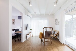 Uma sala de estar moderna com piso de madeira, uma cadeira de madeira, um sofá, uma escrivaninha com cadeira, uma TV fixada na parede e uma janela com cortinas.