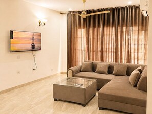 Uma sala de estar com sofá, mesa de centro de vidro, televisão na parede e ventilador de teto.