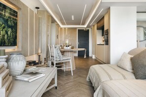 Quarto de hotel moderno com piso de madeira, área de jantar com mesa e cadeiras, cama com colcha listrada e uma grande janela com vista para árvores.