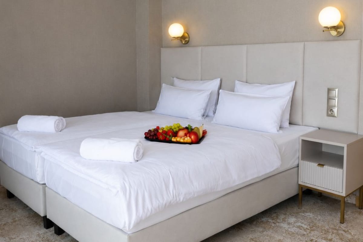 Cama de casal com roupa de cama branca, uma cesta de frutas sobre a cama e uma mesa de cabeceira com abajur.