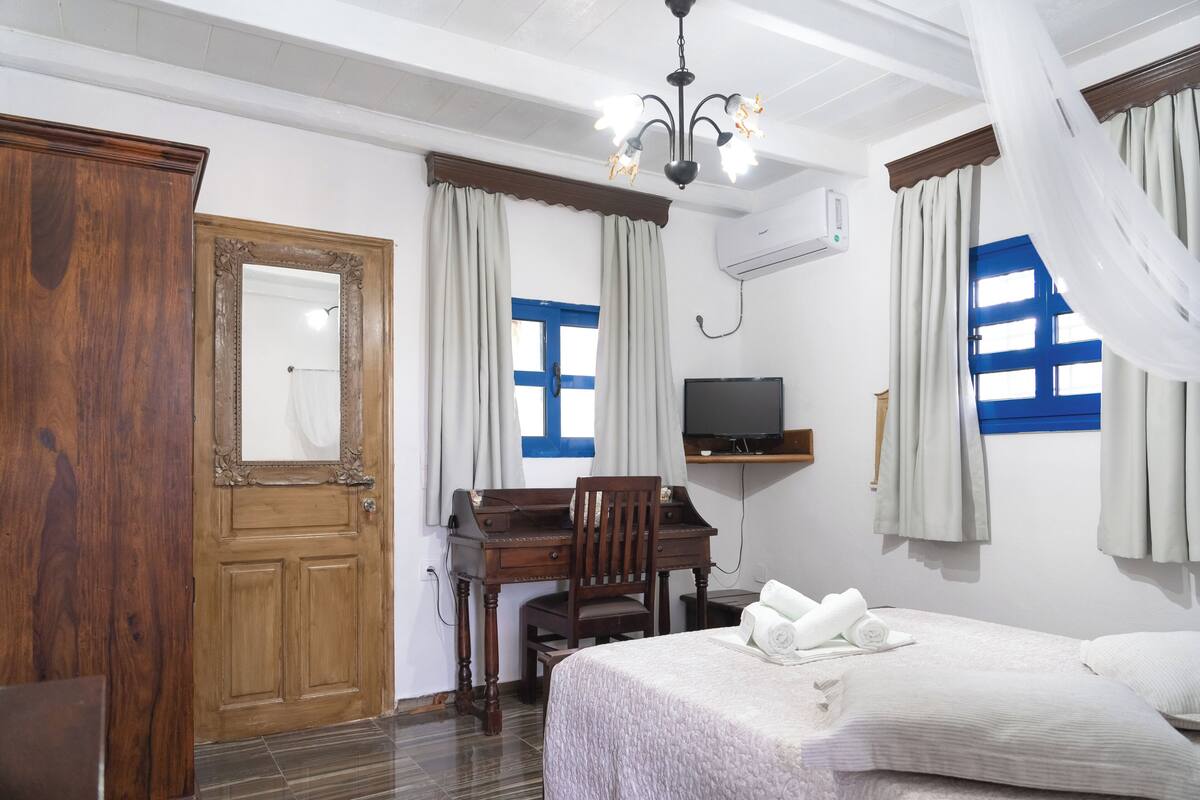 Quarto de hotel com escrivaninha de madeira, cama com toalhas dobradas, televisão e lustre.