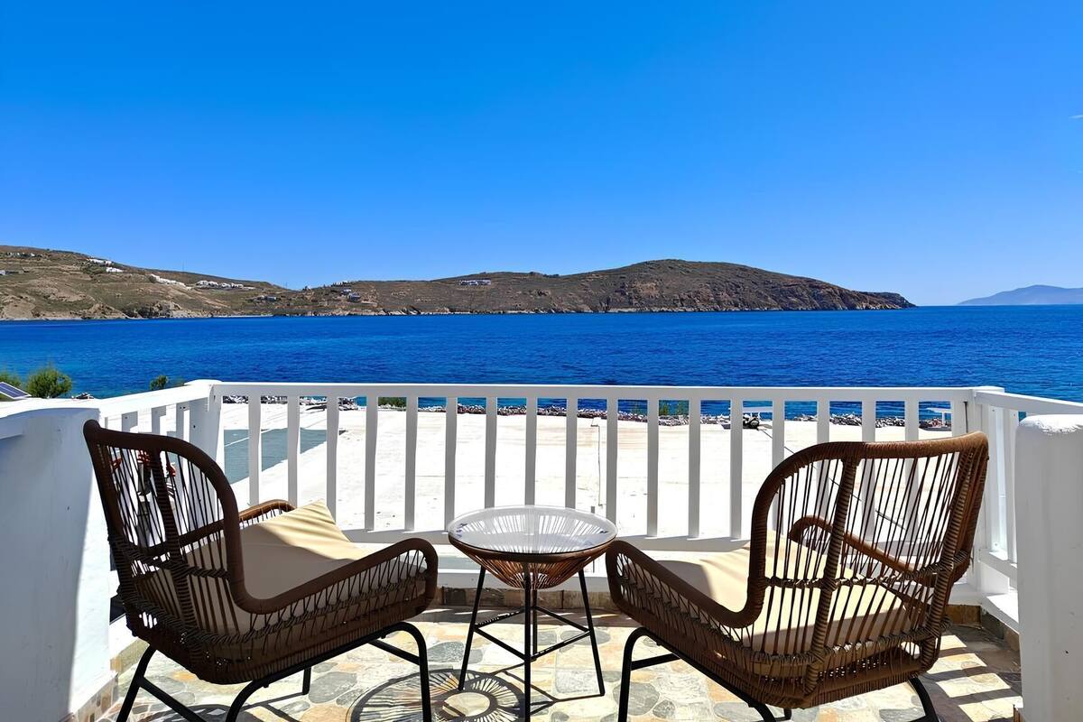 Varanda com duas cadeiras de vime e uma mesa pequena, com vista para o mar azul e colinas distantes.
