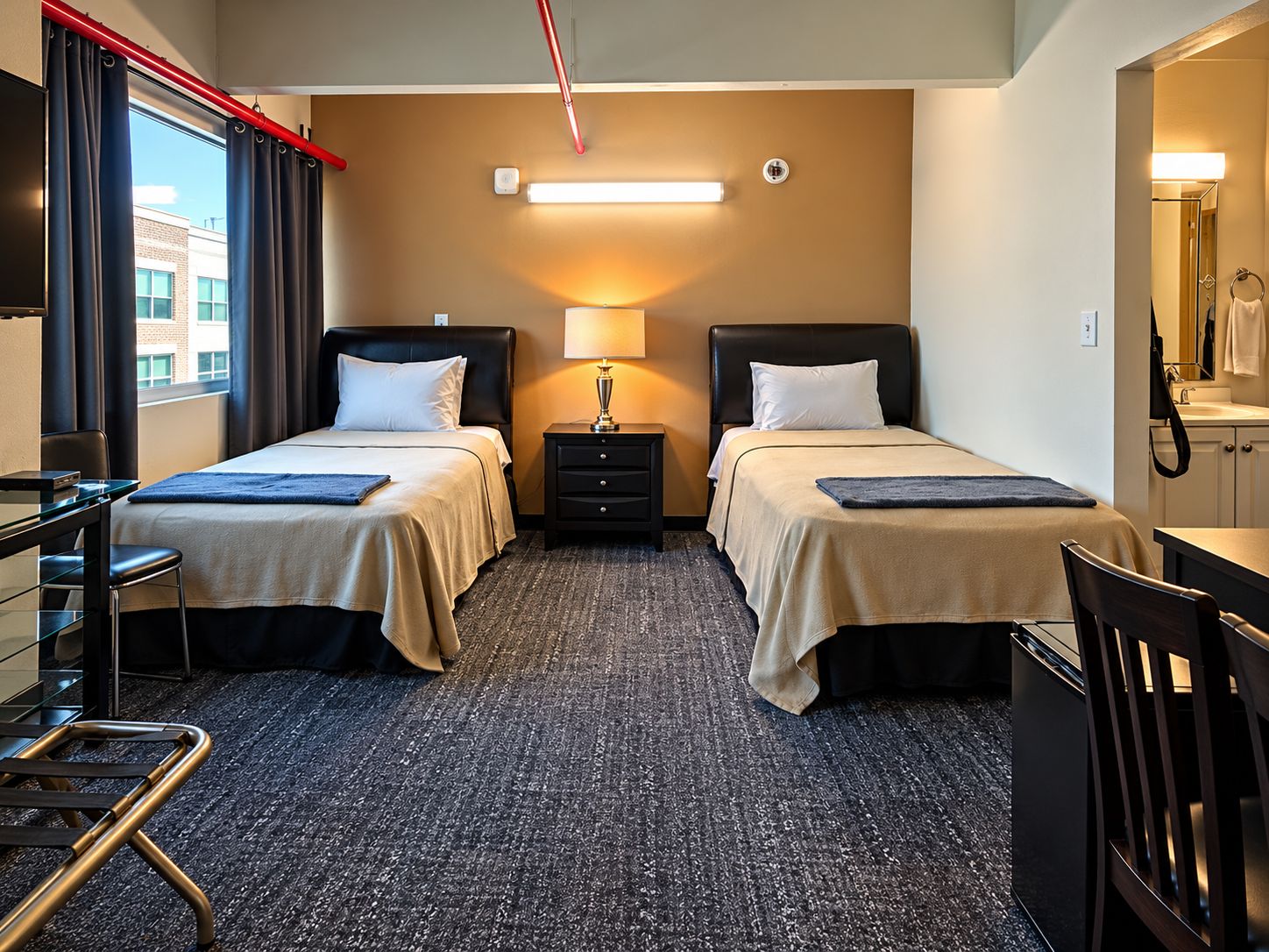 Quarto de hotel com duas camas, uma escrivaninha, uma cadeira, um abajur e vista para um edifício.