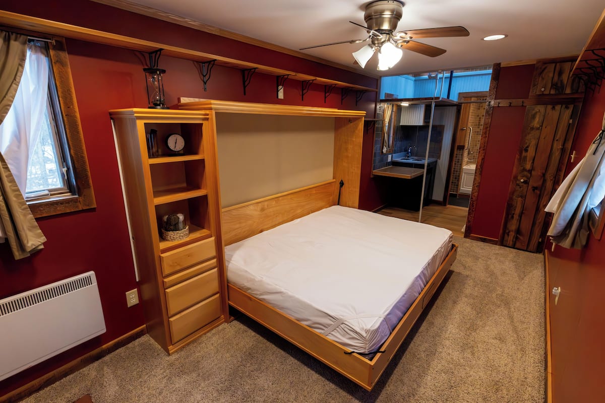 Um quarto com cama de madeira, mesa de cabeceira, estante, radiador e ventoinha no teto.