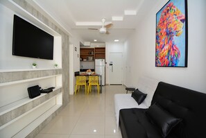 Uma sala de estar com um sofá preto, uma televisão de tela plana, um ventilador de teto e um quadro colorido.